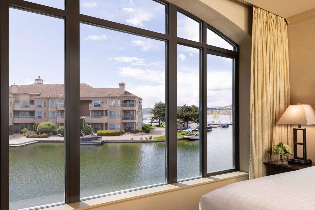 Quarto de hotel com grandes janelas, uma cama, uma mesa de cabeceira, um abajur e vista para um corpo d'água e edifícios.