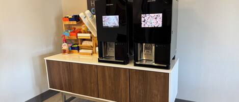 Uma estação de café com duas máquinas de espresso, um dispensador de café e uma variedade de cápsulas de café sobre um balcão de madeira.