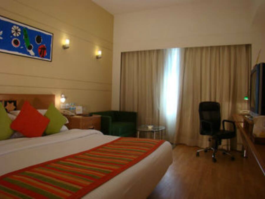 Quarto de hotel com cama, escrivaninha, cadeira, televisão e janela com cortinas.