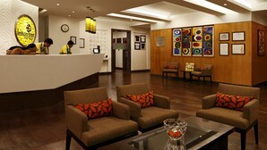 Uma área de recepção de hotel com balcão, dois recepcionistas, um relógio, certificados emoldurados e uma parede com desenhos circulares coloridos.
