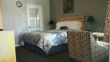Quarto de hotel com uma cama, uma cadeira, uma escrivaninha e um abajur.