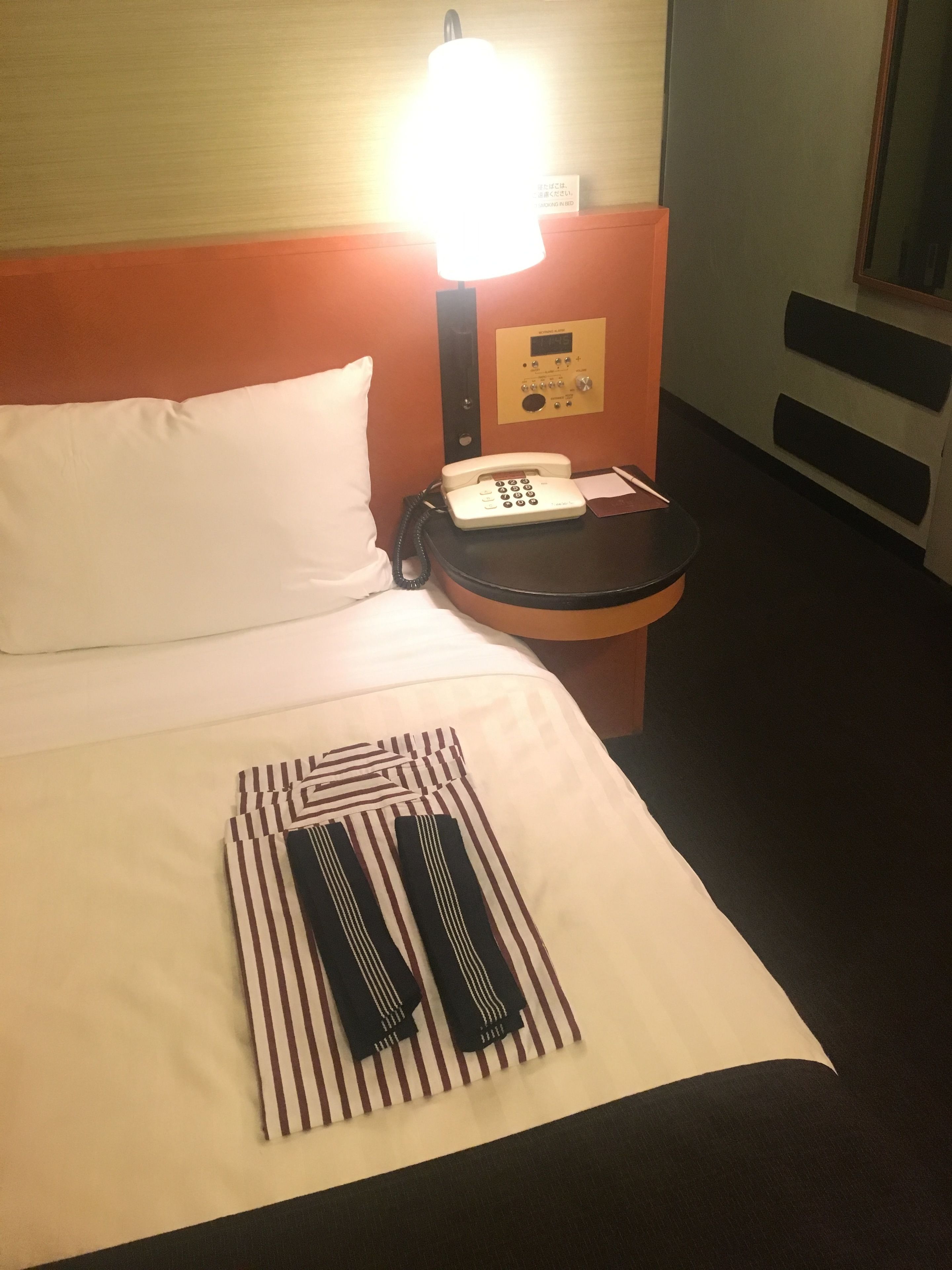 Quarto de hotel com cama, mesa de cabeceira com telefone, luminária e relógio digital.