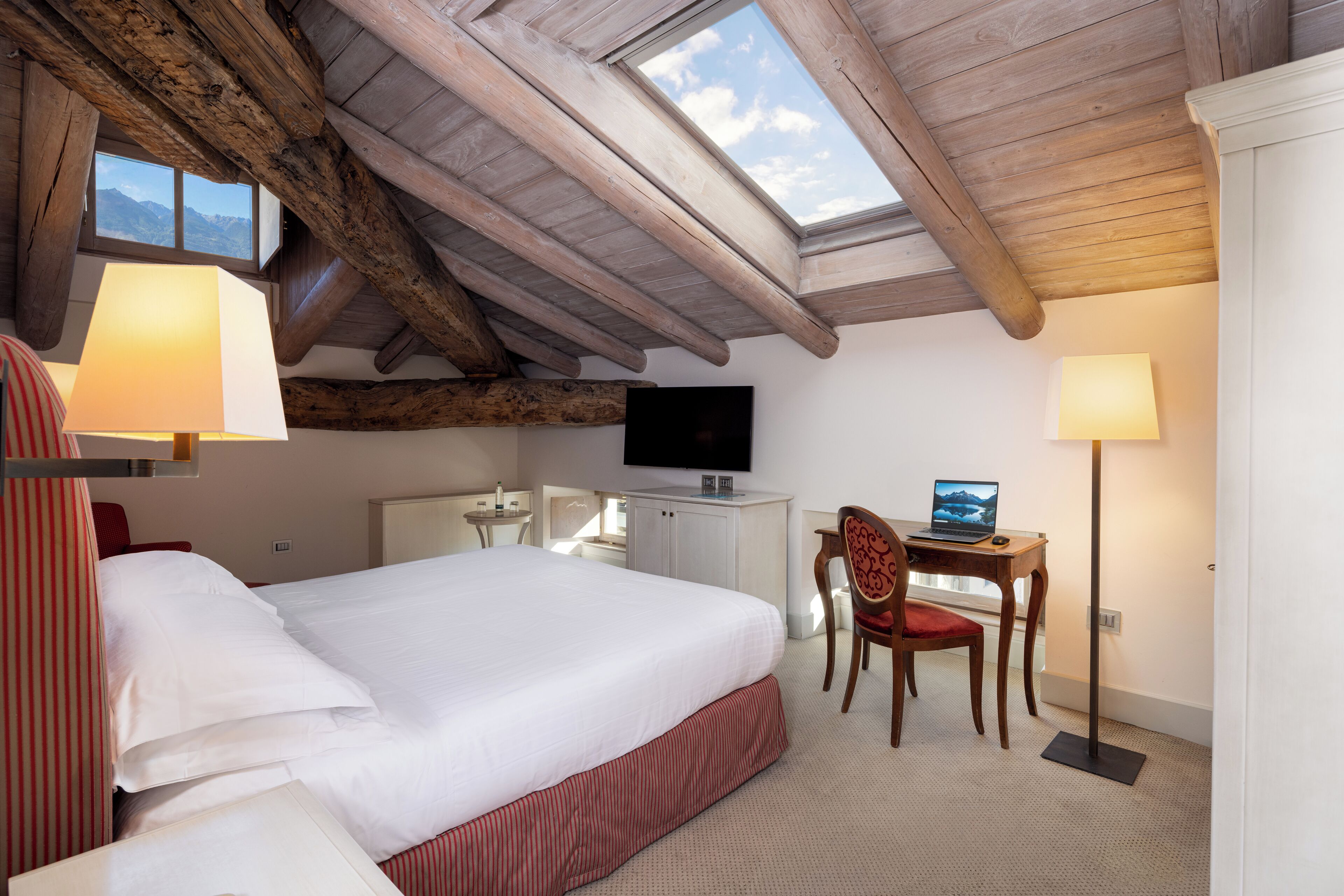Quarto de hotel com uma cama grande, uma escrivaninha com computador, televisão e claraboia.