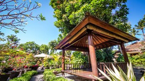 Um gazebo de madeira com telhado de azulejos, cercado por vegetação exuberante e plantas em vasos.
