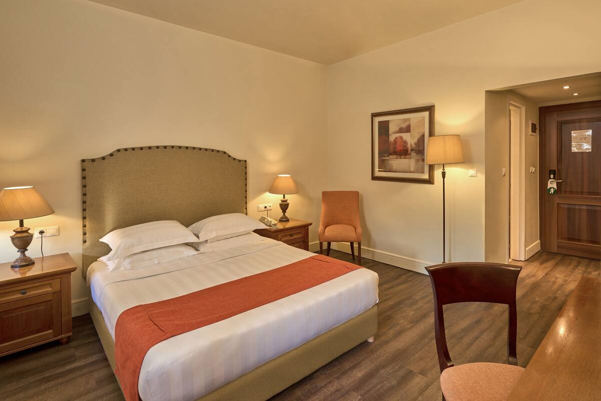 Quarto de hotel com cama, duas mesas de cabeceira com luminárias, uma cadeira, um quadro na parede e uma porta com o logotipo da Starbucks.