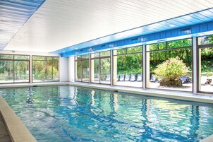 Piscina interior com paredes de vidro transparente, piso ladrilhado e vista para o verde lá fora.
