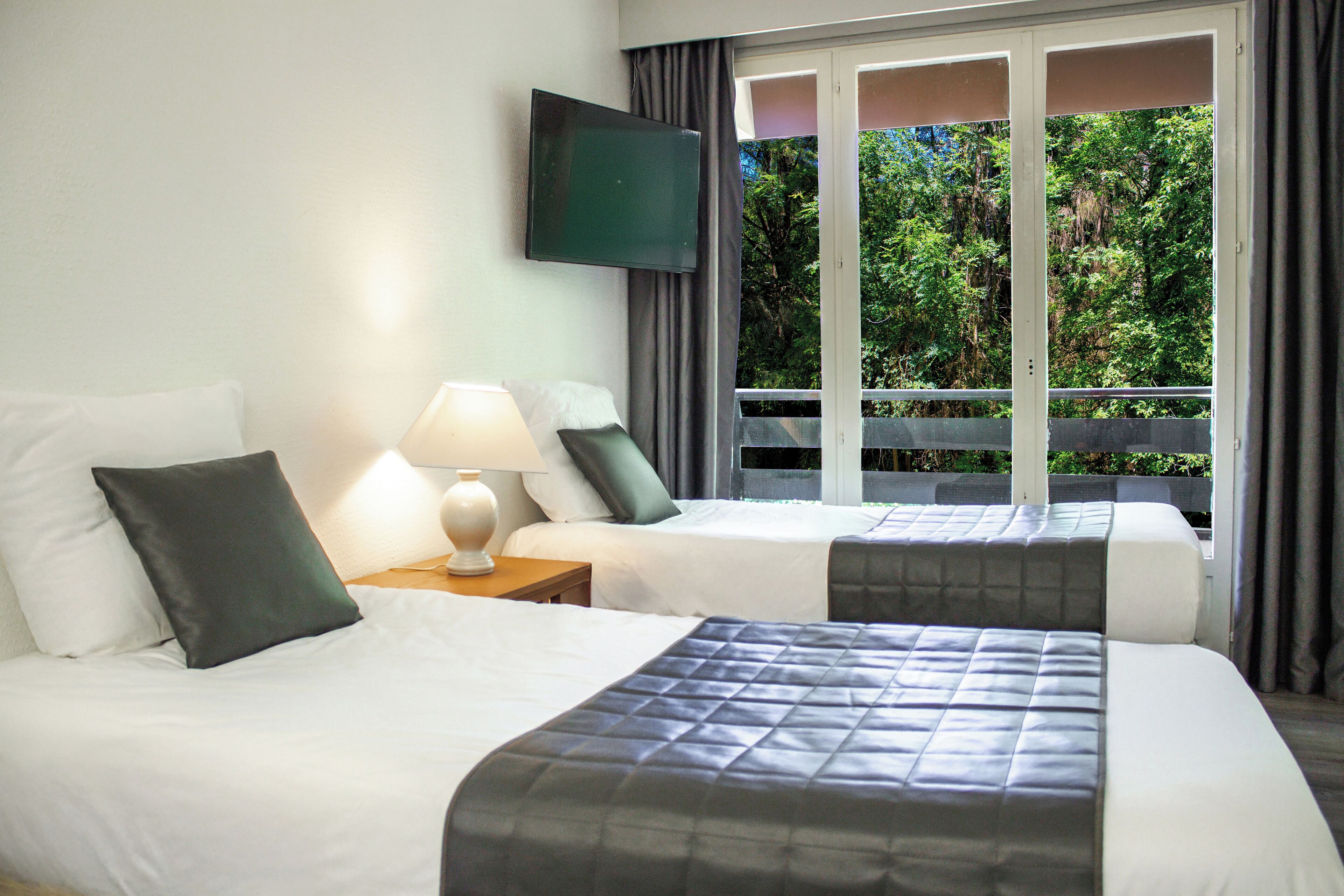 Quarto de hotel com duas camas, uma televisão fixada na parede e uma varanda com vista para o verde.