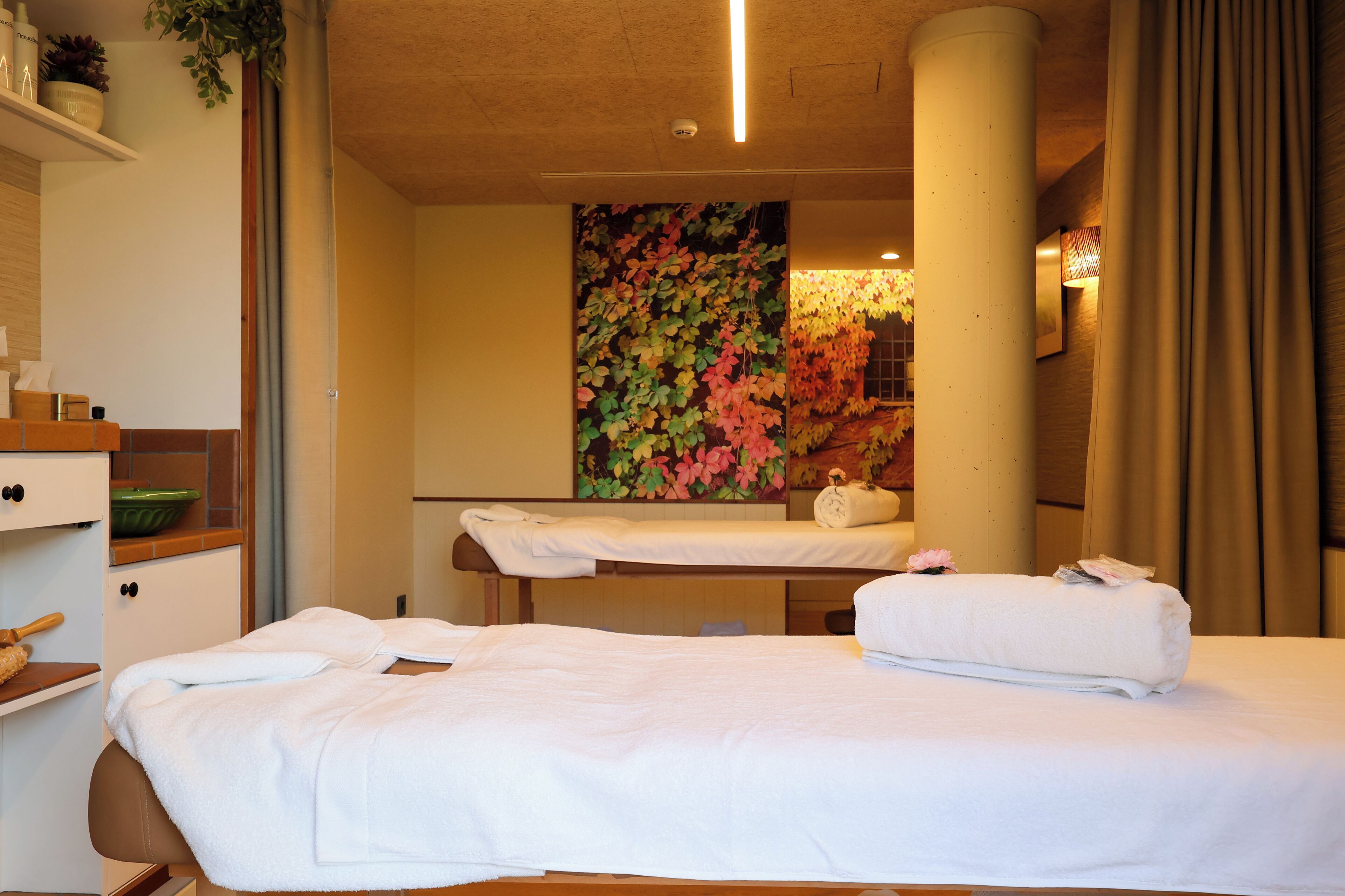 Quarto de hotel com uma cama, mesa de massagem, parede com um mural floral, prateleira com amenities e janela com cortinas.