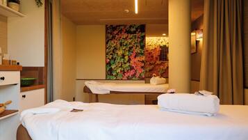 Quarto de hotel com uma cama, mesa de massagem, parede com um mural floral, prateleira com amenities e janela com cortinas.