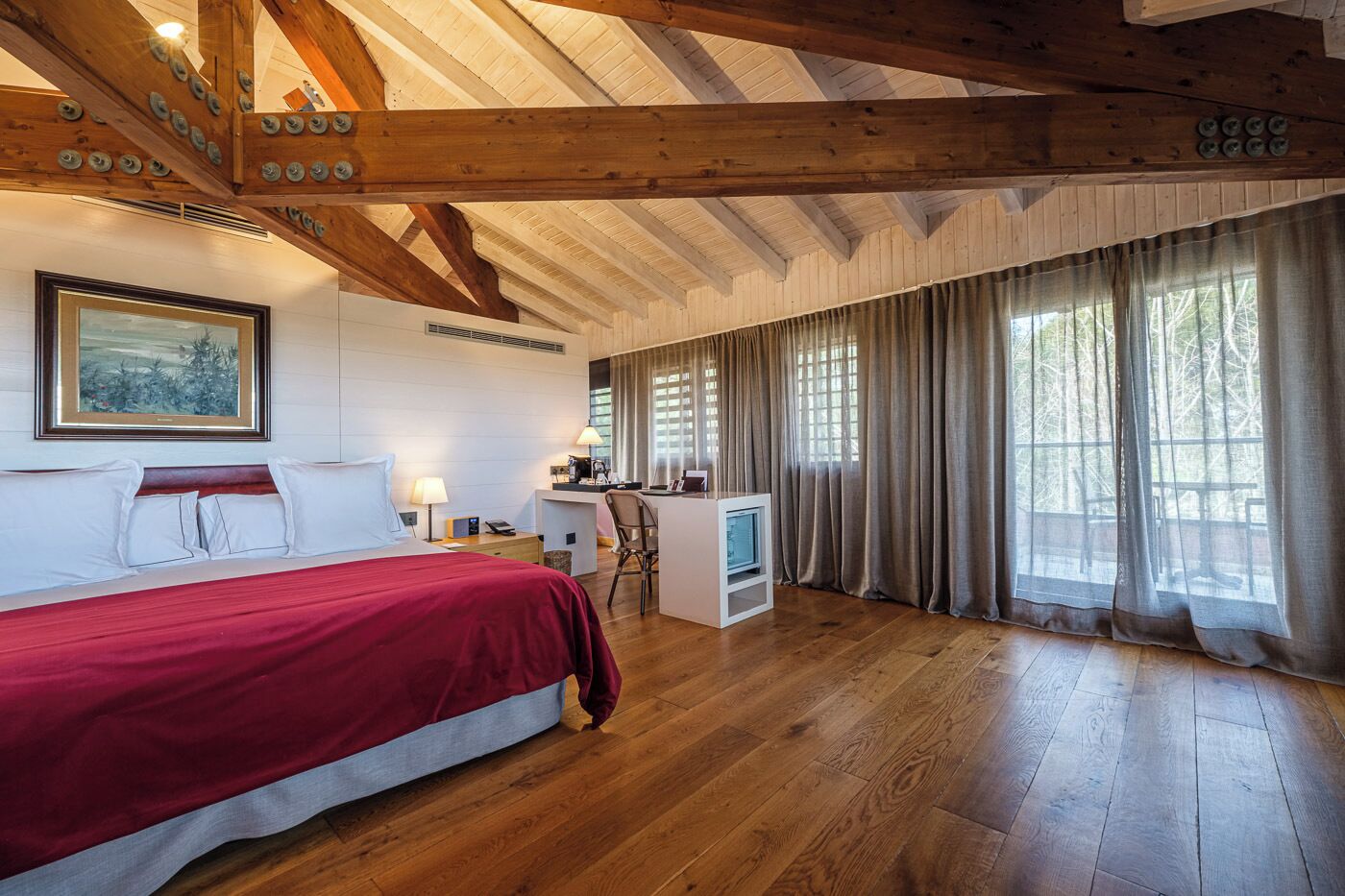 Quarto de hotel com teto de madeira, cama com colcha vermelha, escrivaninha com computador e janelas amplas com cortinas.