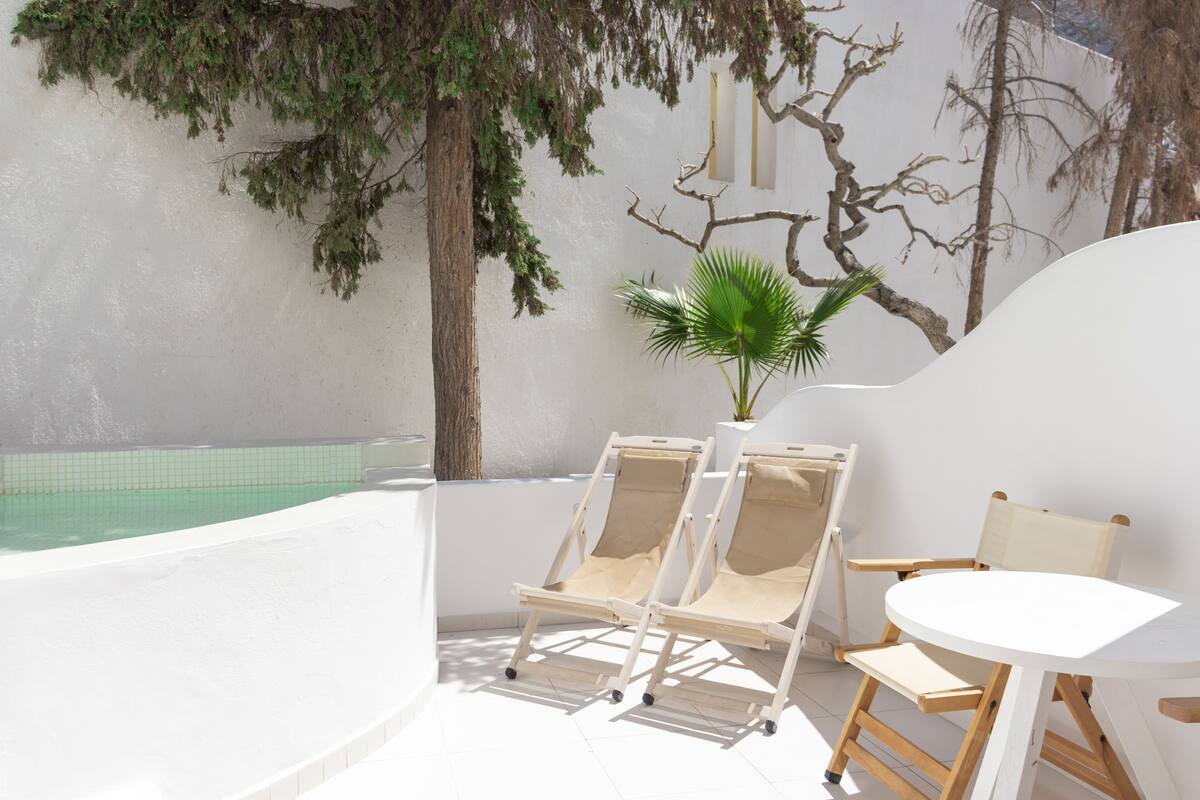 Uma área externa ensolarada com assentos, duas poltronas de descanso, uma mesinha, um vaso de planta e uma piscina.