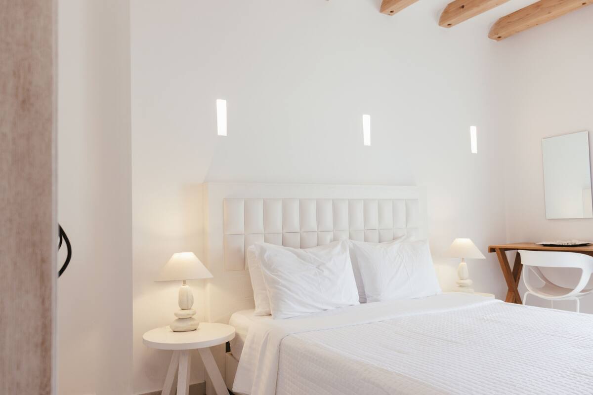 Quarto de hotel moderno com uma cama grande, duas mesas de cabeceira com luminárias, uma escrivaninha de madeira e uma cadeira.