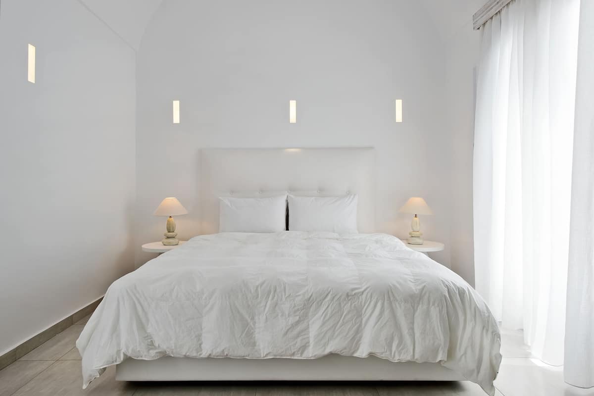 Quarto minimalista com uma cama grande, duas mesas de cabeceira com luminárias e uma parede branca lisa.