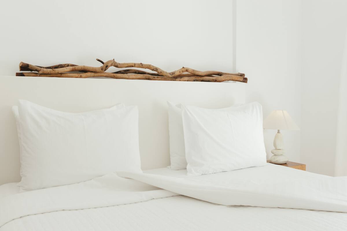 Uma cama bem arrumada com lençóis e almofadas brancas, uma cabeceira de madeira com detalhes em madeira natural e uma mesa de cabeceira com abajur.
