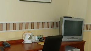Quarto de hotel compacto com uma secretária de madeira, um pequeno frigorífico, uma cadeira preta, telefone, máquina de café e um quadro na parede.