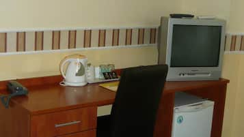 Quarto de hotel compacto com uma secretária de madeira, um pequeno frigorífico, uma cadeira preta, telefone, máquina de café e um quadro na parede.