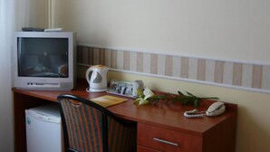 Quarto de hotel compacto com secretária de madeira, pequeno frigorífico, cadeira, telefone, chaleira e televisão.