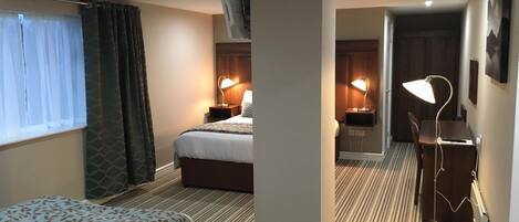 Quarto de hotel com cama, escrivaninha e cadeira. Possui duas mesas de cabeceira com abajures. O quarto tem uma janela com cortinas.