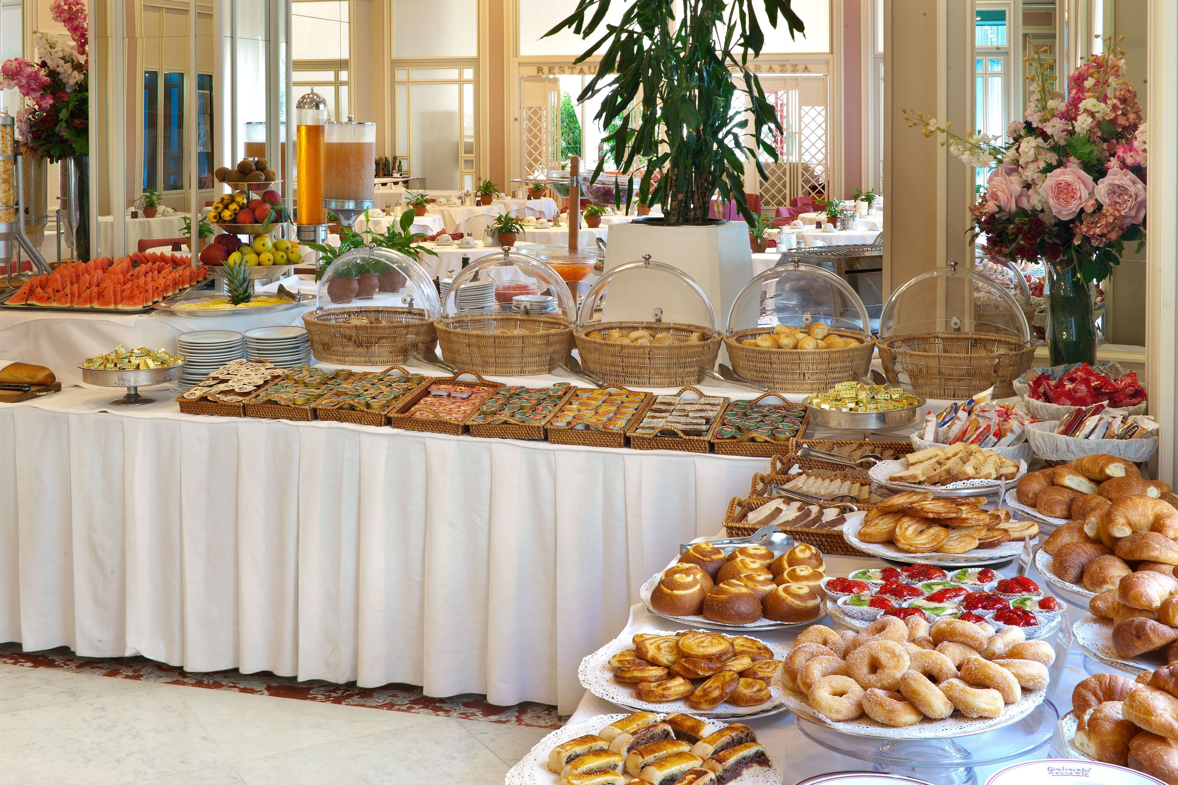 Uma mesa de buffet com diversos alimentos, incluindo doces, frutas e pão, cobertos por cúpulas transparentes.