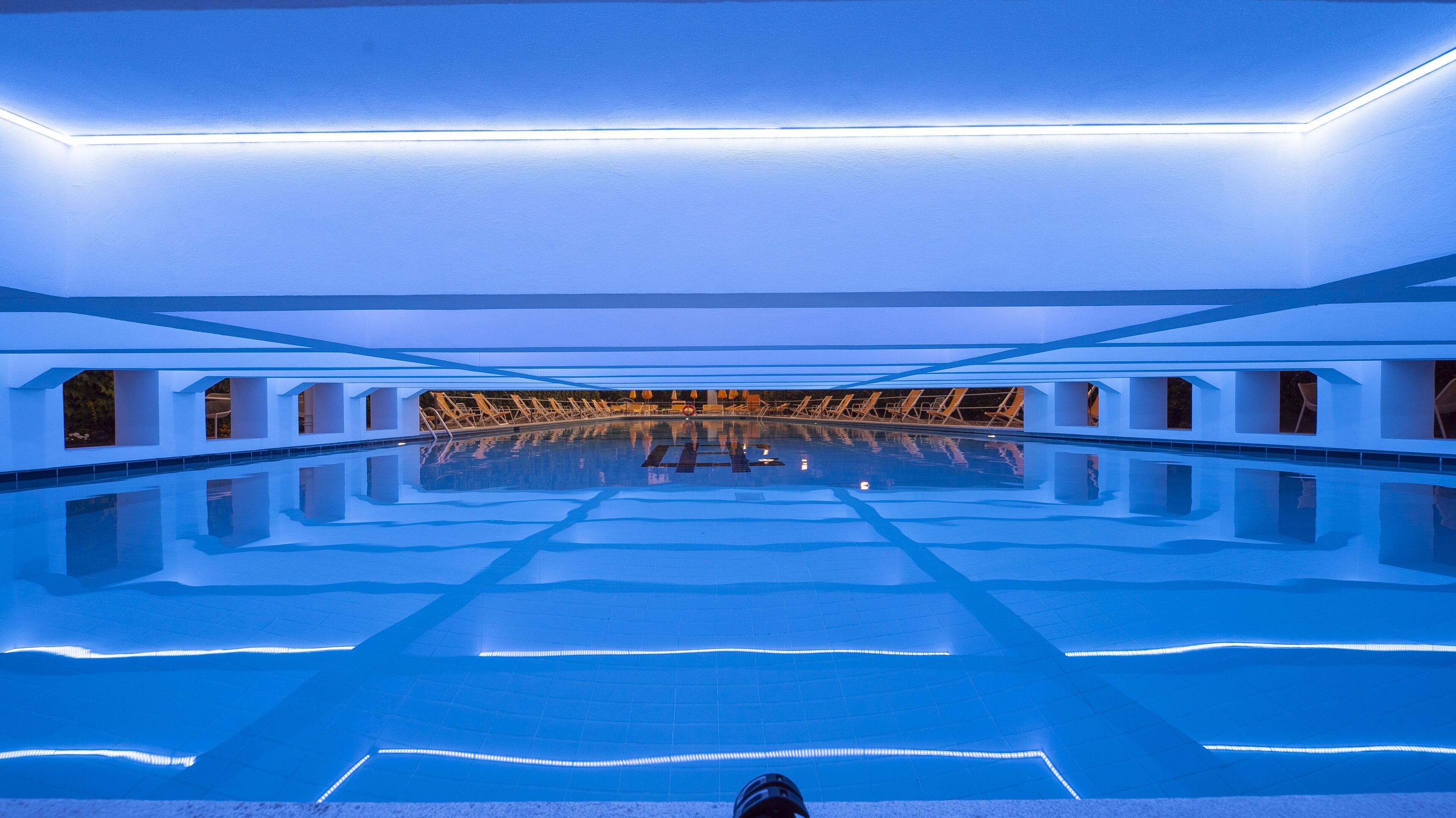 Uma piscina interior ampla com água azul cristalina, rodeada por paredes brancas e várias janelas.