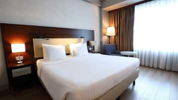 Quarto de hotel com uma cama grande, duas mesas de cabeceira com luminárias, uma cadeira, uma pequena mesa redonda e vista para o exterior através de cortinas.