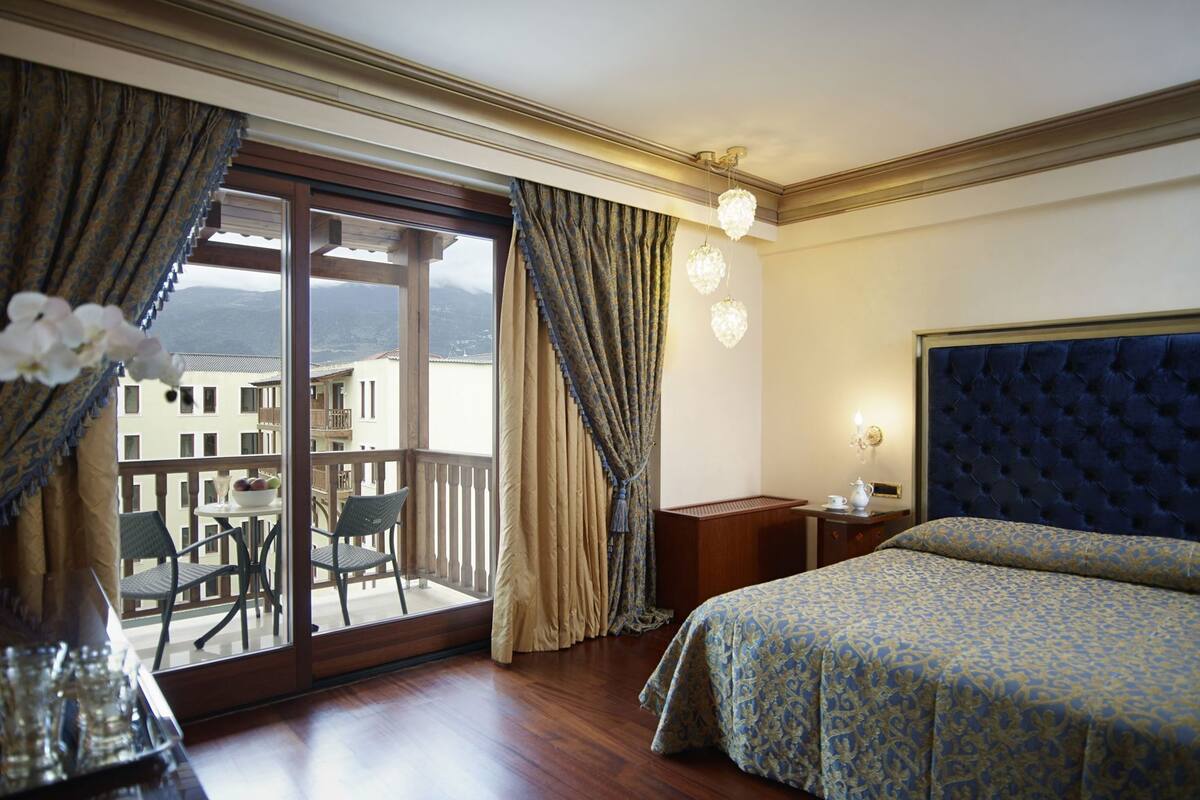 Quarto de hotel com varanda, cama com colcha estampada, mesa de cabeceira com abajur e vista para edifícios.