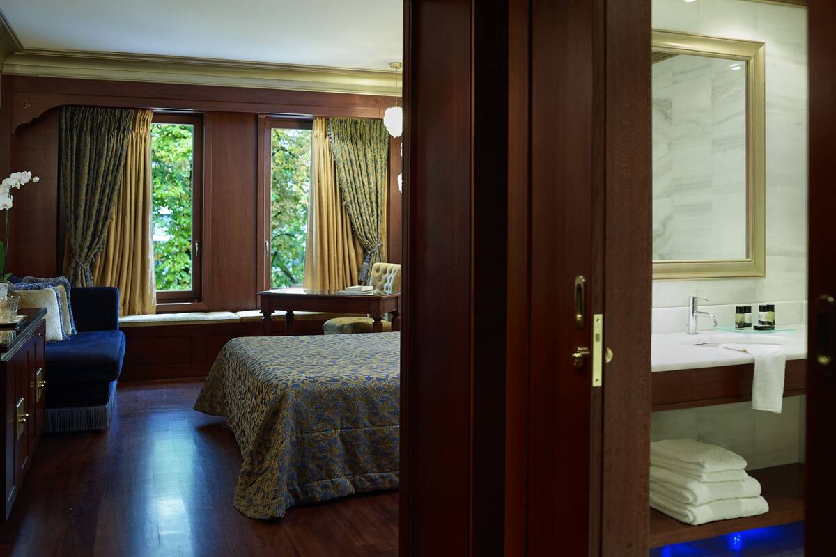 Quarto de hotel com cama, escrivaninha, cadeira, janela com cortinas, banheiro com espelho e pia, e vista para o verde lá fora.