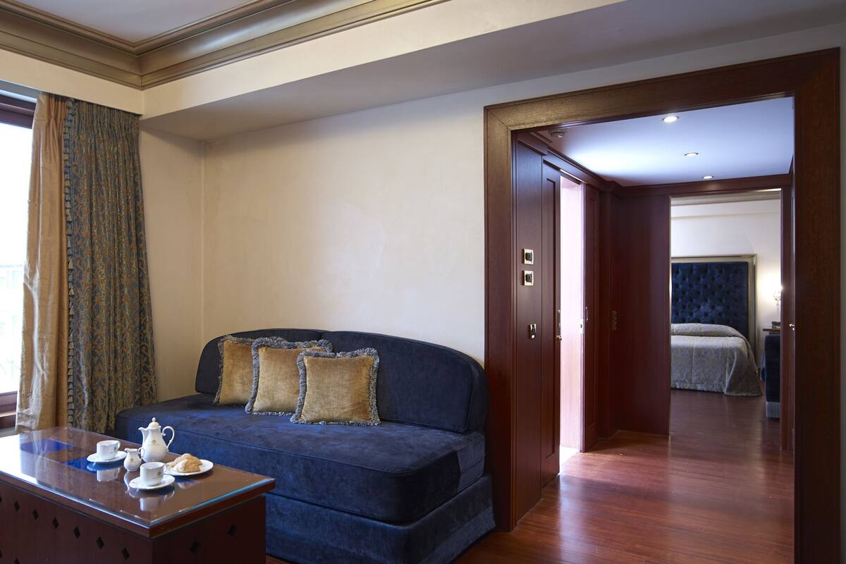 Quarto de hotel com sofá azul, mesa de centro de madeira e cama com cabeceira azul.