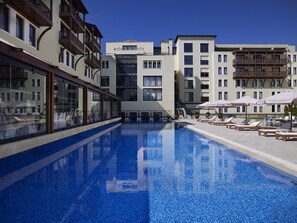 Piscina com espreguiçadeiras e guarda-sóis, cercada por edifícios altos com varandas.