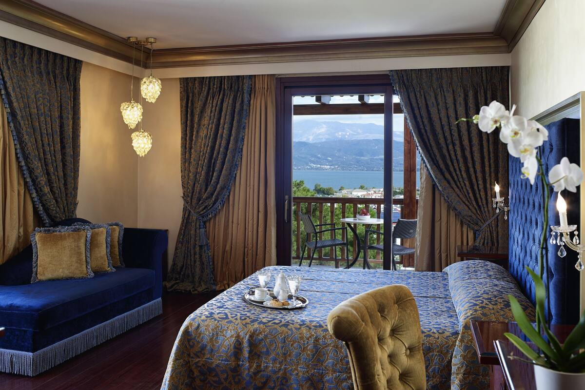 Quarto de hotel com cama, sofá, mesa de jantar e vista para um lago.