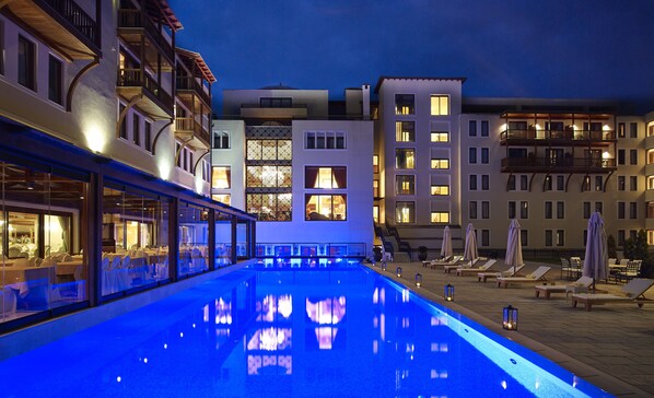 Piscina com espreguiçadeiras e guarda-sóis, cercada por edifícios altos com varandas e janelas iluminadas.