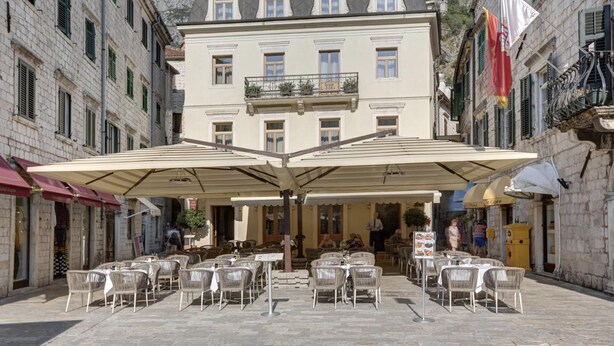 Um café ao ar livre com mesas e cadeiras, um edifício com varanda e bandeiras.