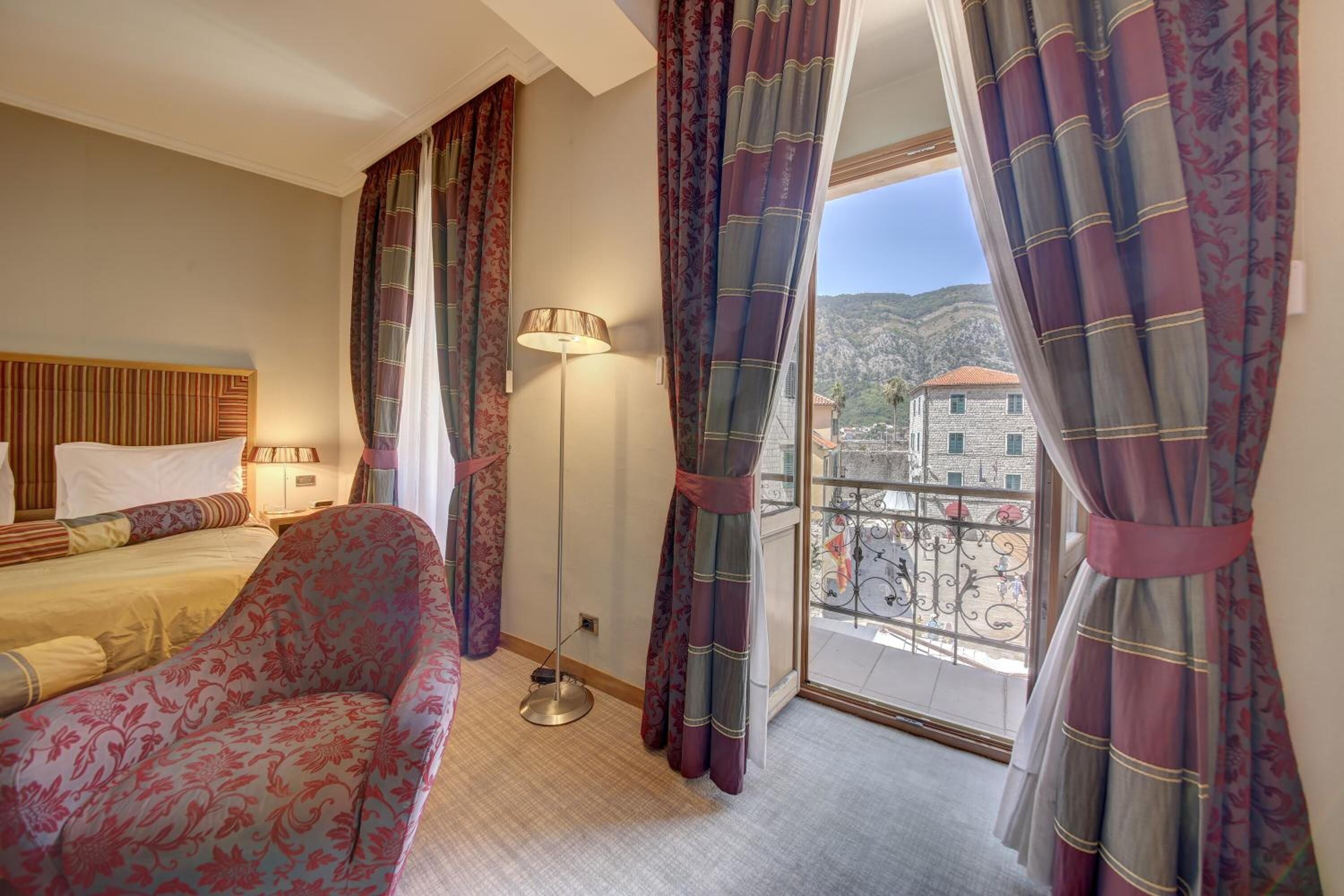 Quarto de hotel com cama, uma cadeira, abajur e varanda com vista para as colinas.