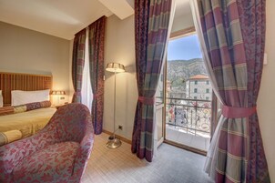 Quarto de hotel com cama, uma cadeira, abajur e varanda com vista para as colinas.