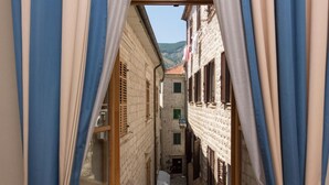 Vista de uma rua estreita através de uma janela com cortinas, com edifícios de pedra e janelas com persianas, sob um céu claro.