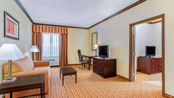 Quarto de hotel com sofá, escrivaninha com televisão, cadeira, cômoda e janela com cortinas.