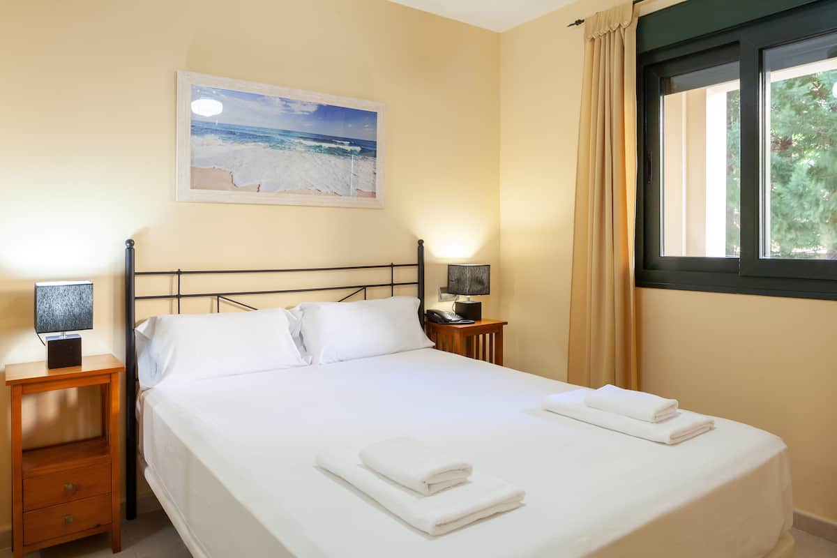 Uma cama bem arrumada com lençóis brancos e duas toalhas dobradas, uma mesa de cabeceira com abajur, e uma pintura de uma praia na parede.