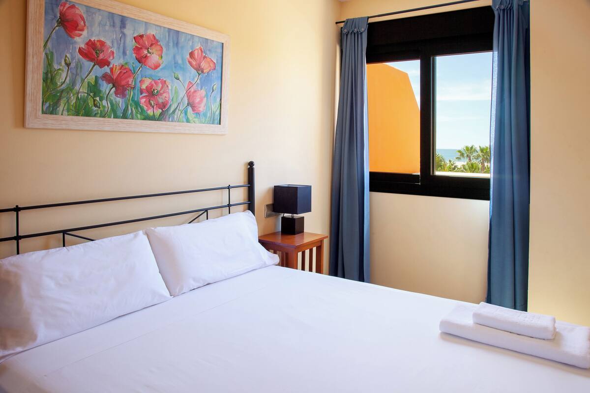 Uma cama bem arrumada com lençóis brancos, um quadro de flores vermelhas na parede, uma mesa de cabeceira com abajur e uma janela com cortinas.