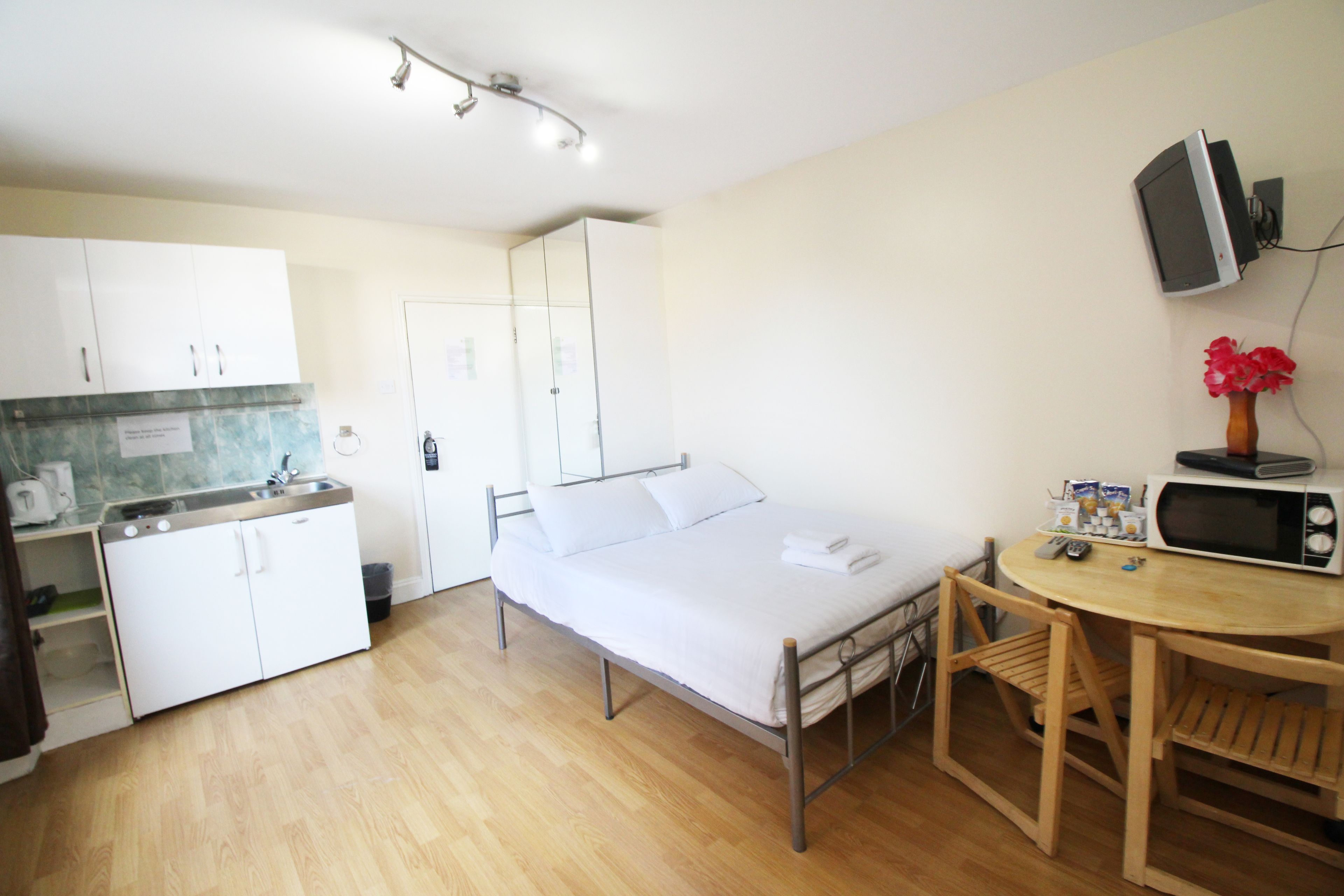 Quarto compacto com cama, pequeno espaço de cozinha, mesa de jantar e televisão.