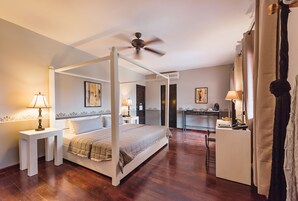 Uma cama com dossel e cortina, uma escrivaninha com luminária e uma cadeira em quarto de hotel com piso de madeira e ventilador de teto.