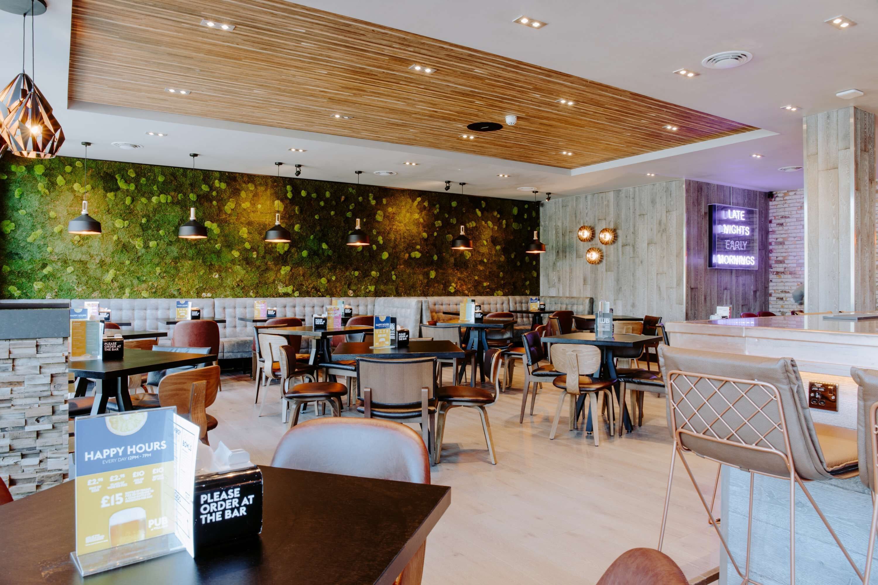 Um restaurante moderno com painéis de teto de madeira, parede coberta de folhagem verde e várias mesas e cadeiras.