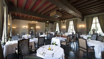 Uma sala de jantar com várias mesas preparadas para hóspedes, com vigas de madeira clássicas e lustres.