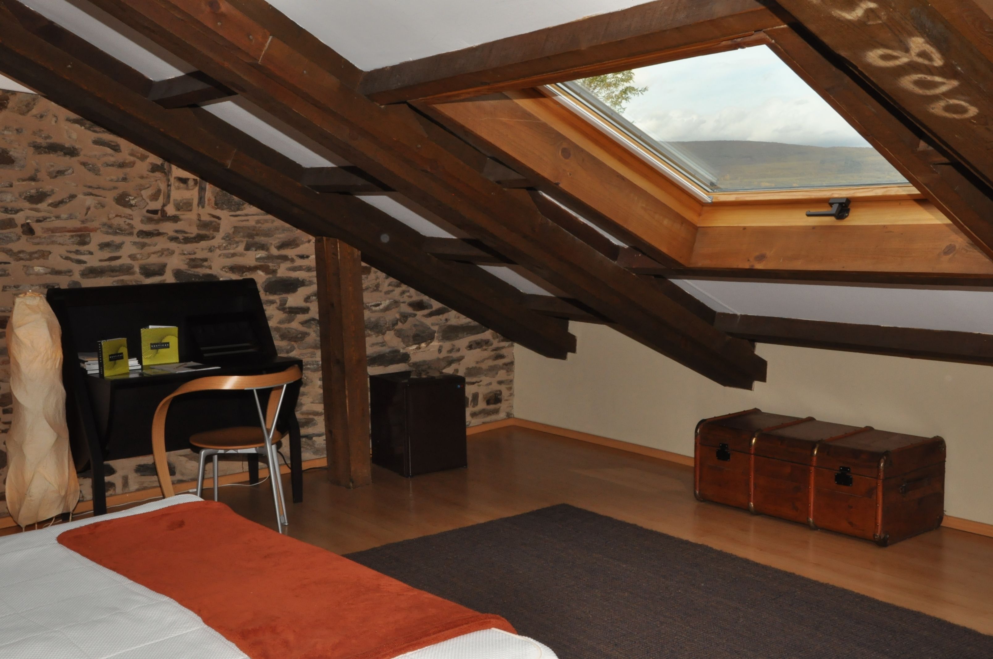 Um quarto com teto inclinado, claraboia, escrivaninha com cadeira, cama, mala e parede de pedra.