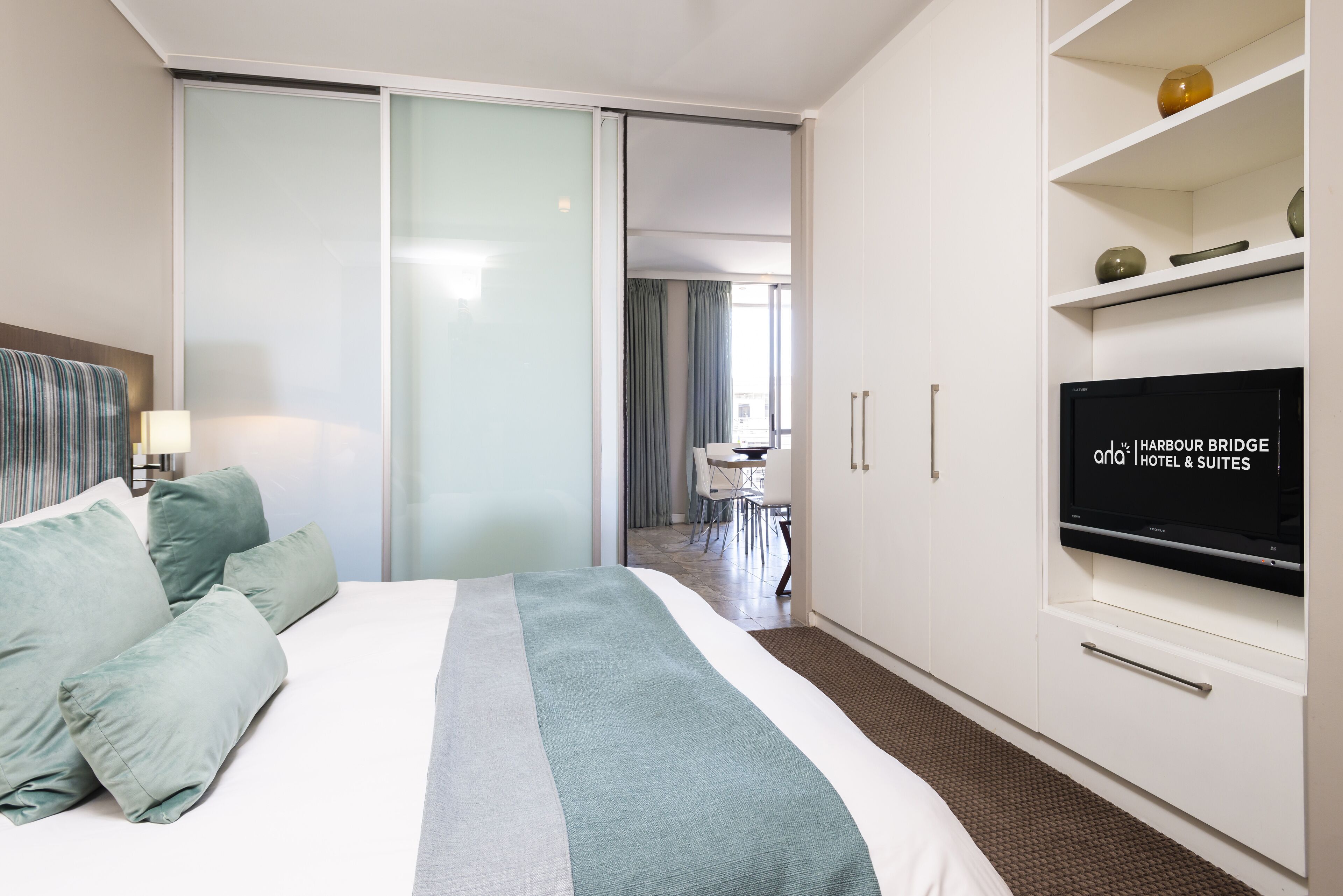 Quarto de hotel com cama, televisão, porta deslizante e vista para a sala de jantar.