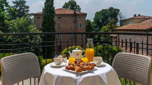 Mesa de varanda posta com croissants, café e sumo de laranja, com vista para um pátio com árvores e edifícios.