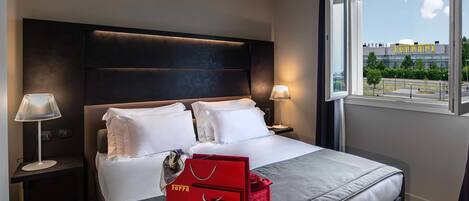 Quarto de hotel moderno com uma cama grande, duas mesas de cabeceira com luminárias, uma janela com vista para um prédio e sacolas de compras vermelhas sobre a cama.