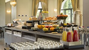 Buffet com diversos alimentos e bebidas, um canto para café e uma grande janela com cortinas.