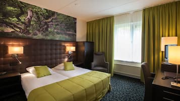 Quarto de hotel com uma cama grande, duas luminárias de cabeceira, uma cadeira, uma escrivaninha com abajur e um quadro de uma floresta na parede.