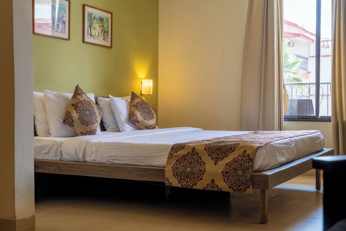 Quarto de hotel com cama, mesa de cabeceira, luminária e janela com cortinas.