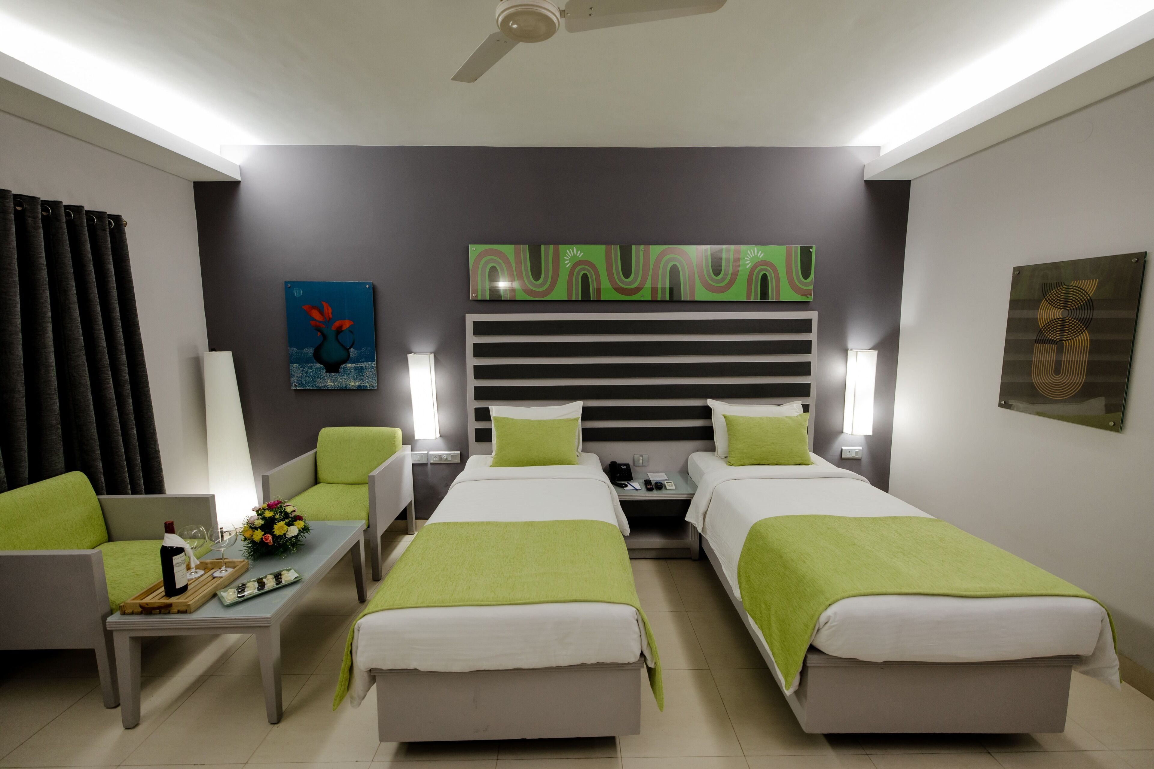 Quarto de hotel moderno com duas camas de solteiro, uma mesa de escritório, uma cadeira, uma pequena mesa com um vaso de flores e um ventilador de teto.